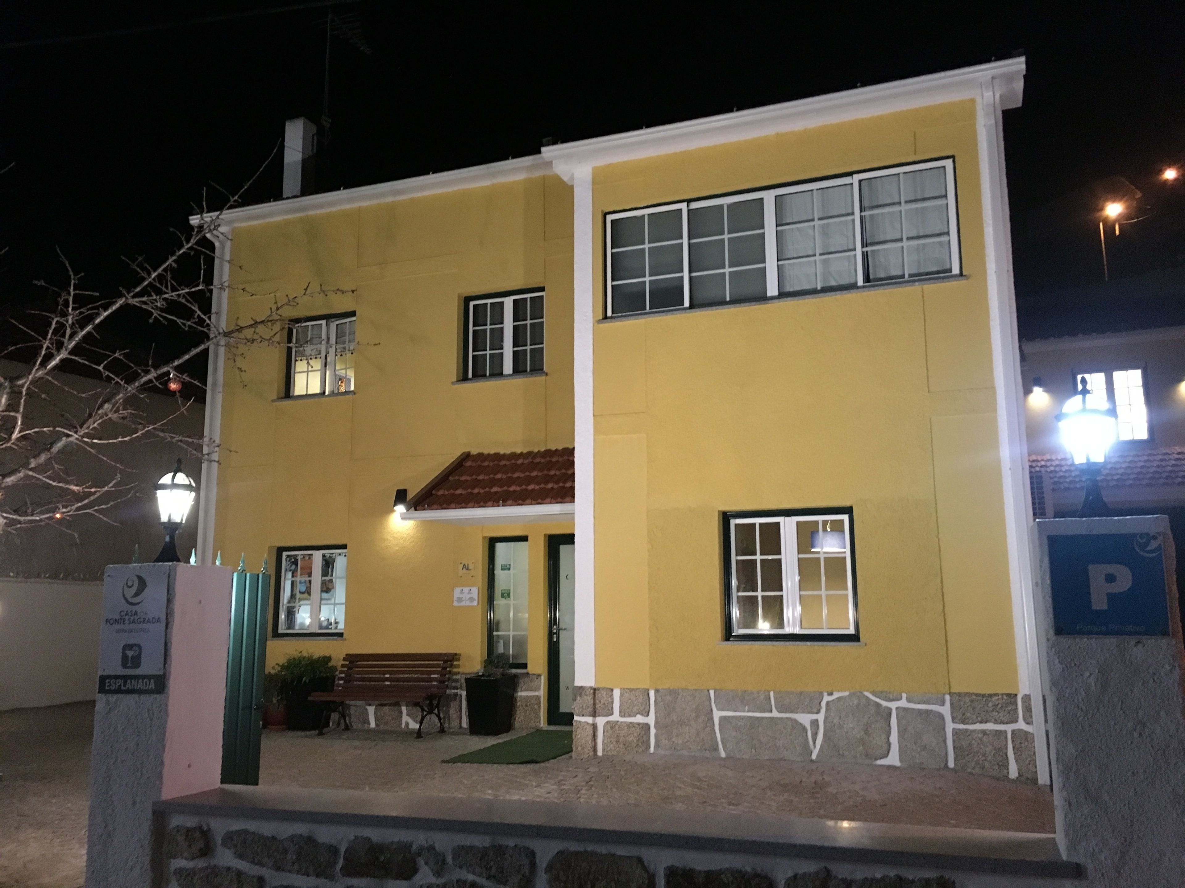 Um edifício de dois andares com fachada amarela, várias janelas, um banco na frente e uma placa indicando área de estacionamento.