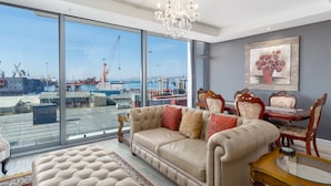 Ampla sala de estar com uma grande janela com vista para um porto, um sofá bege macio, um lustre e uma área de jantar com móveis de madeira.