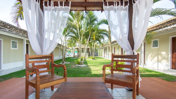 Um banco e mesa de madeira sob um toldo com cortinas brancas, cercado por um jardim com palmeiras e um pátio com ladrilhos.