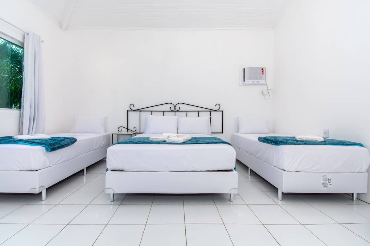 Quarto com três camas brancas, janela com cortinas e ar condicionado fixado na parede.