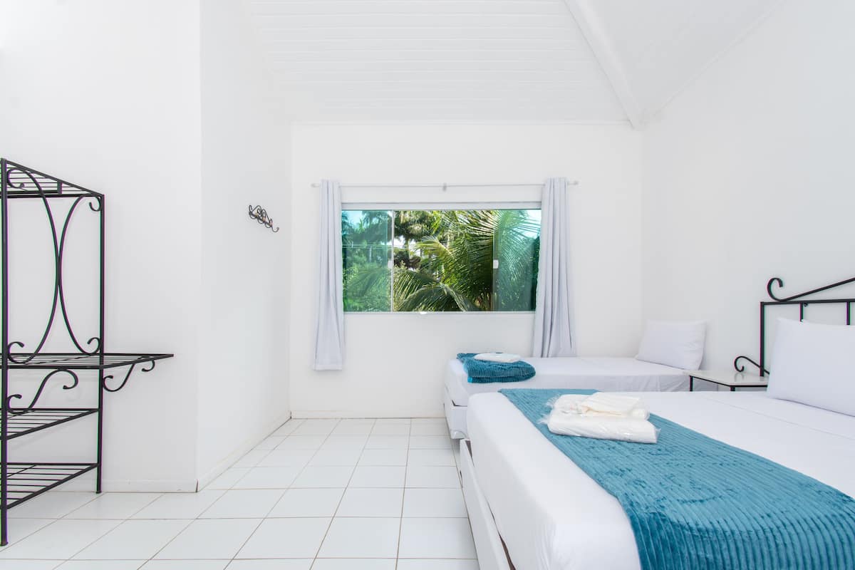 Quarto de hotel com duas camas, janela com cortinas, prateleira de metal e vista para o verde lá fora.