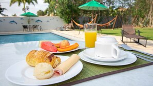 Mesa de café da manhã ao lado de uma piscina, com croissants, frutas, um copo de suco de laranja e uma xícara branca.