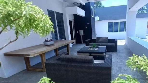 Uma área de estar moderna ao ar livre, com mesa de madeira e sofás com almofadas pretas, rodeada por vasos com plantas.