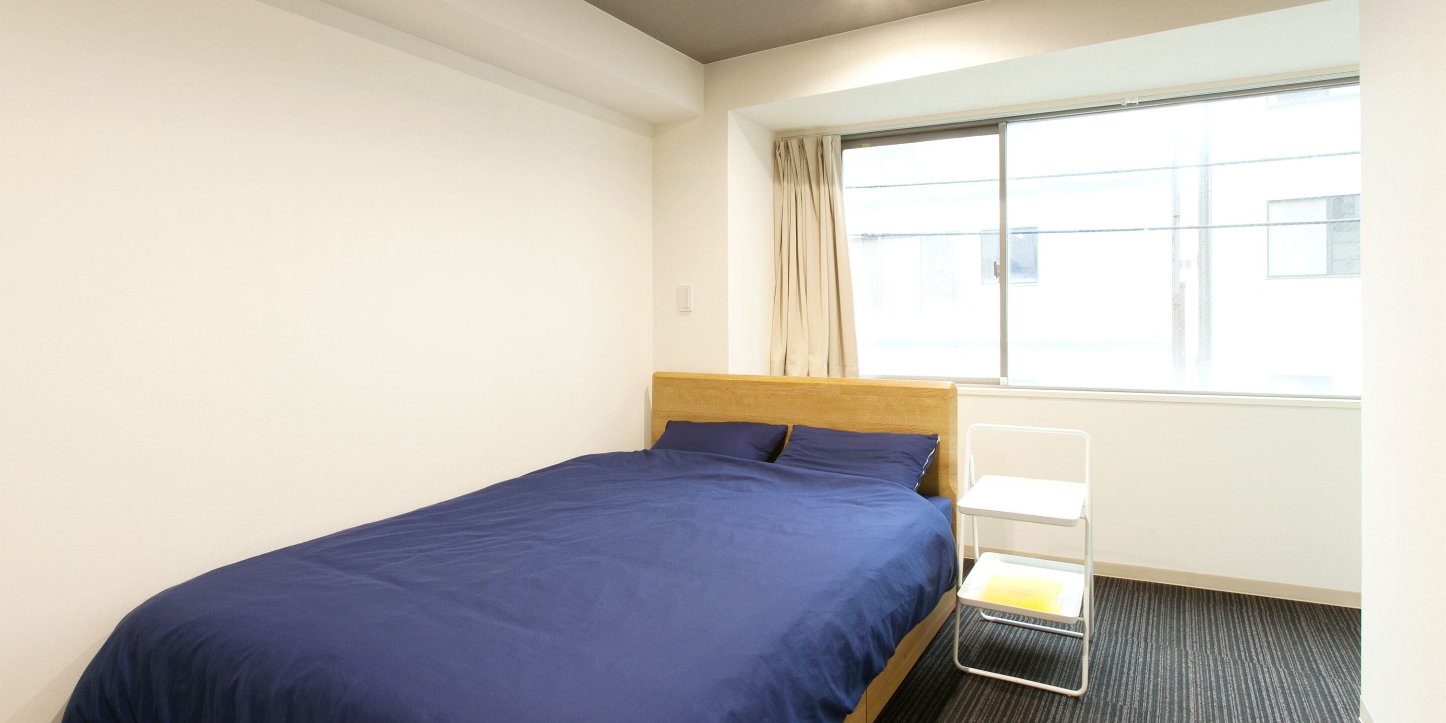 Uma cama de solteiro com cabeceira de madeira em quarto com uma janela grande e uma pequena mesa branca.