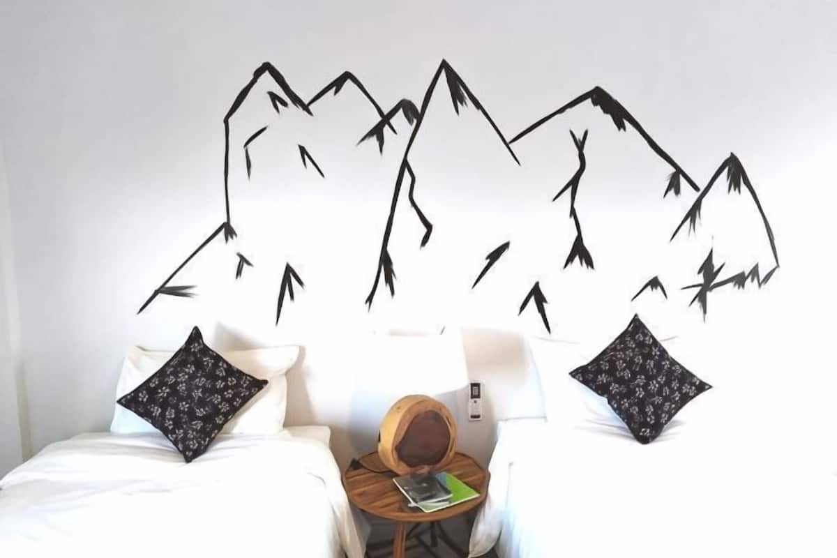 Quarto de hotel com cama, duas almofadas, uma pequena mesa redonda com um livro e um chapéu, e uma parede com desenho de montanha.