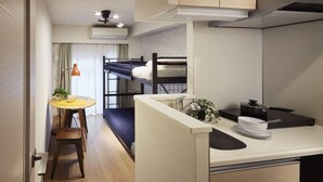 Quarto de hotel compacto com beliches, uma kitchenette pequena, uma mesa de jantar com cadeiras e um vaso de planta.
