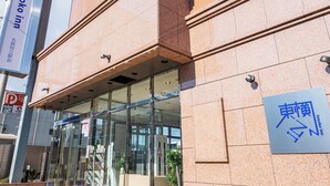 Um edifício moderno com entrada de vidro, sinalização com 'toyoko inn' e placa indicando disponibilidade de estacionamento 24 horas.