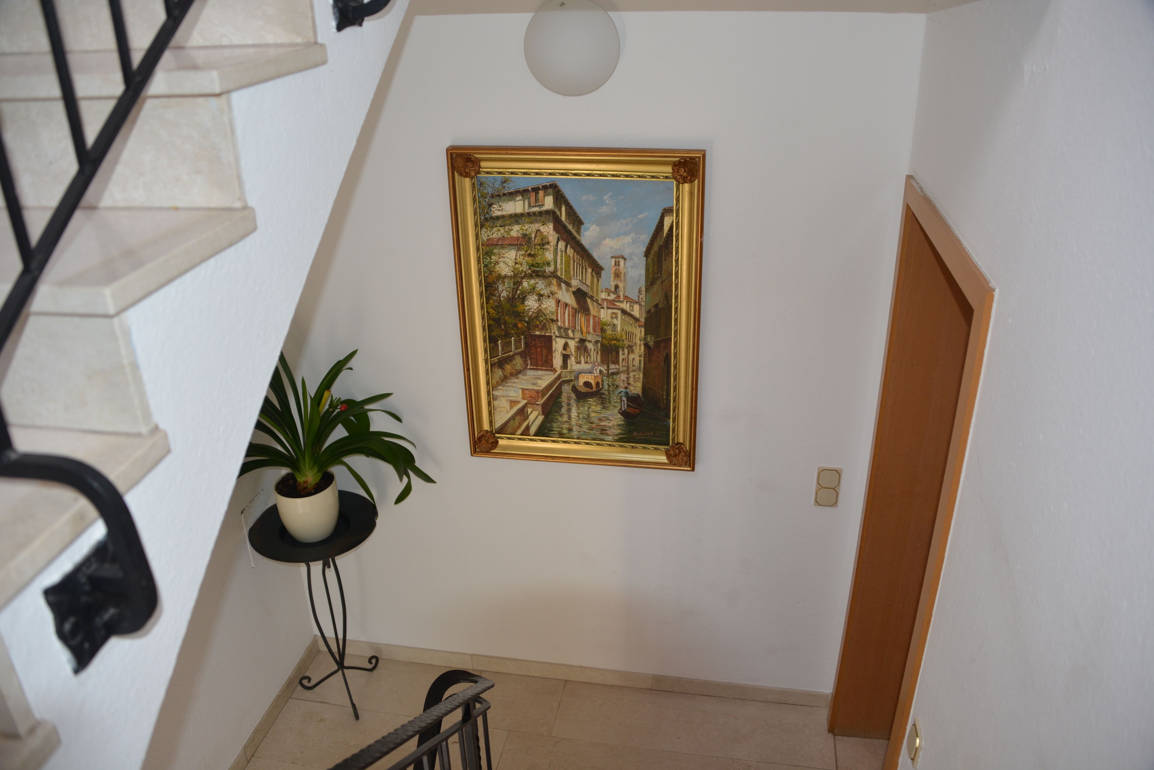 Uma escadaria com um vaso de planta no patamar, um quadro na parede e uma porta à direita.