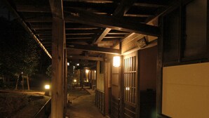 Um corredor pouco iluminado, com vigas de madeira e piso de concreto, que leva a um edifício com janelas protegidas por grades.