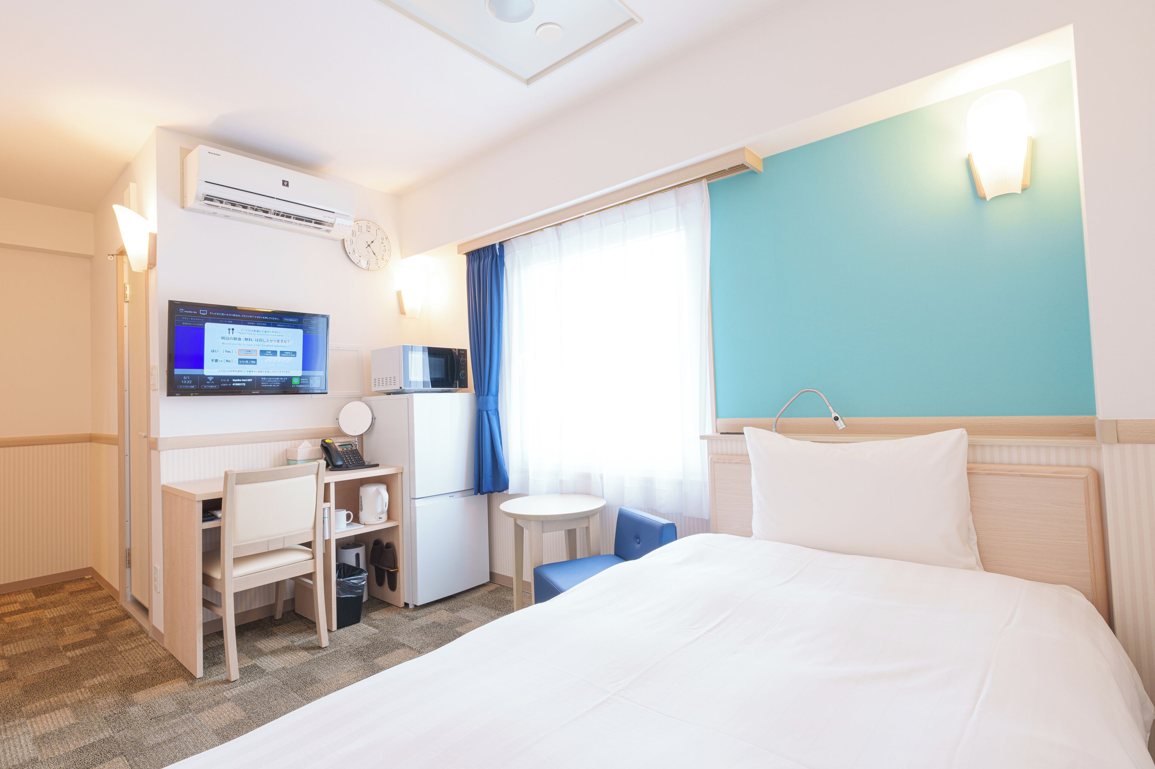 Quarto de hotel com uma cama, uma escrivaninha com computador, um micro-ondas e uma pequena mesa.