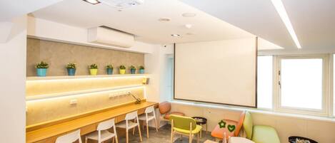 Uma sala de reuniões moderna com uma mesa comprida, cadeiras brancas, uma tela de projetor e plantas em vasos.