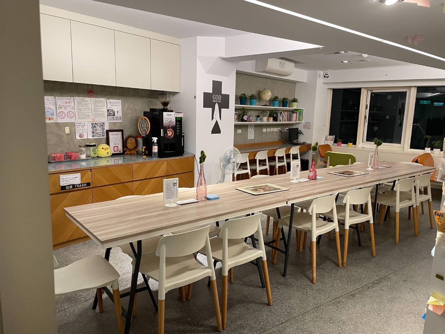 Uma sala de reunião moderna com uma mesa comprida, cadeiras brancas, uma máquina de café e uma estante com plantas e livros.