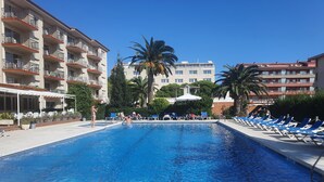 Piscina com espreguiçadeiras e área de estar coberta, cercada por edifícios altos e palmeiras.
