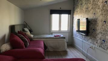 Quarto com cama, sofá vermelho, TV fixada na parede e janela com vista para o exterior.