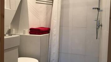 Casa de banho pequena e moderna, com sanita, chuveiro e uma toalha branca no chão.