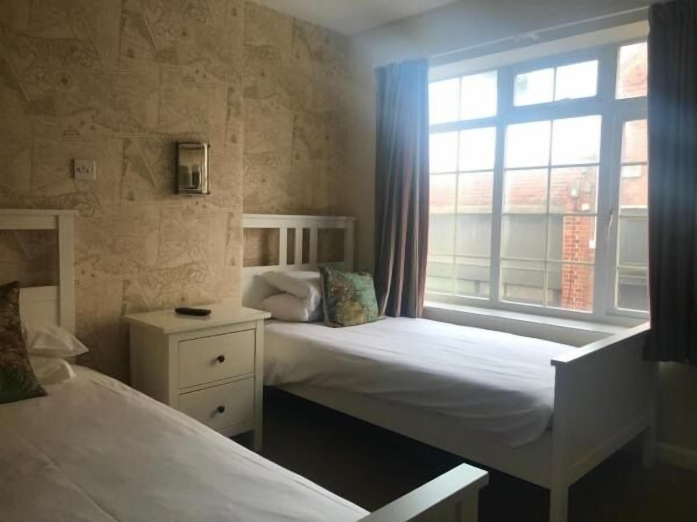 Quarto com duas camas, uma pequena mesa de cabeceira com gavetas, um criado-mudo e uma janela com cortinas.