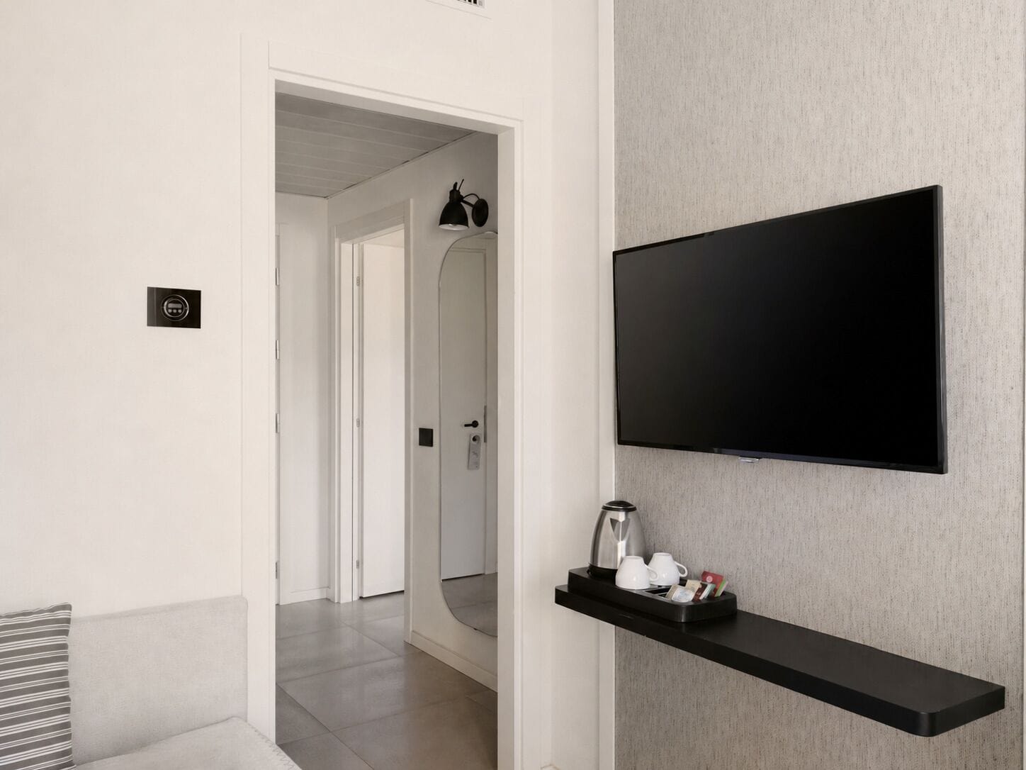 Quarto de hotel moderno com televisão de tela plana na parede, uma prateleira preta com chaleira e xícaras, e uma porta que dá para outro cômodo.