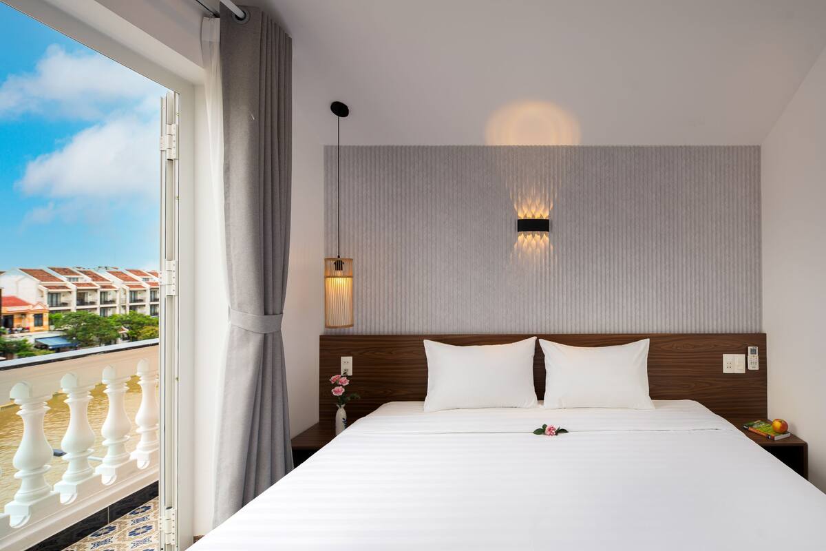 Quarto de hotel com uma cama grande, uma mesa de cabeceira com abajur, vista para edifícios e uma janela com cortina.