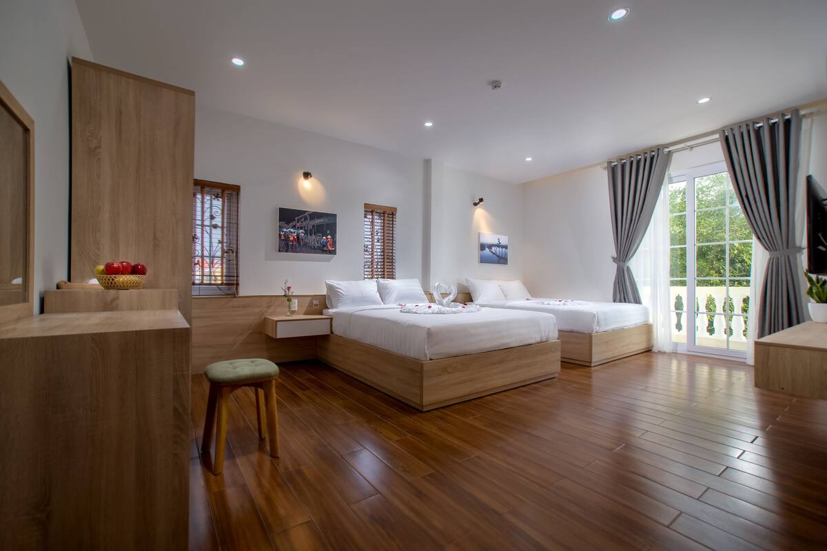 Quarto de hotel moderno com piso de madeira, duas camas, uma mesa de escritório e uma varanda com cortinas.
