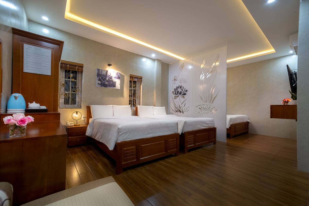Quarto de hotel moderno com piso de madeira, duas camas, uma mesa com chaleira azul e televisão na parede.
