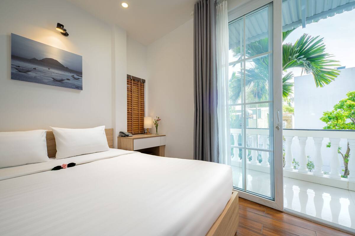Quarto de hotel com uma cama grande, uma mesa de cabeceira com luminária, um quadro na parede e uma varanda com vista para palmeiras.