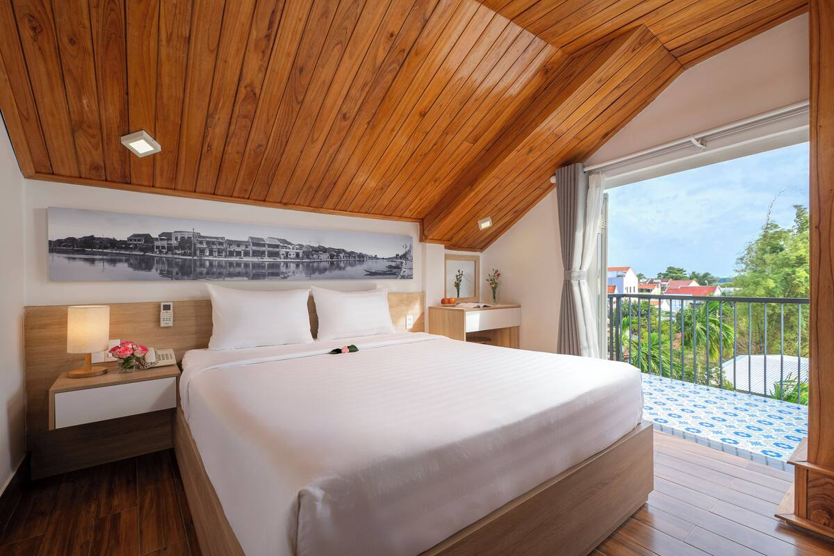 Quarto de hotel com teto de madeira, uma cama grande, mesa de cabeceira com abajur, criado-mudo com telefone e vista para a varanda externa.