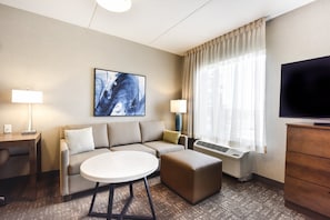 Quarto de hotel com sofá, banquinho, mesa redonda, televisão e obra de arte na parede.