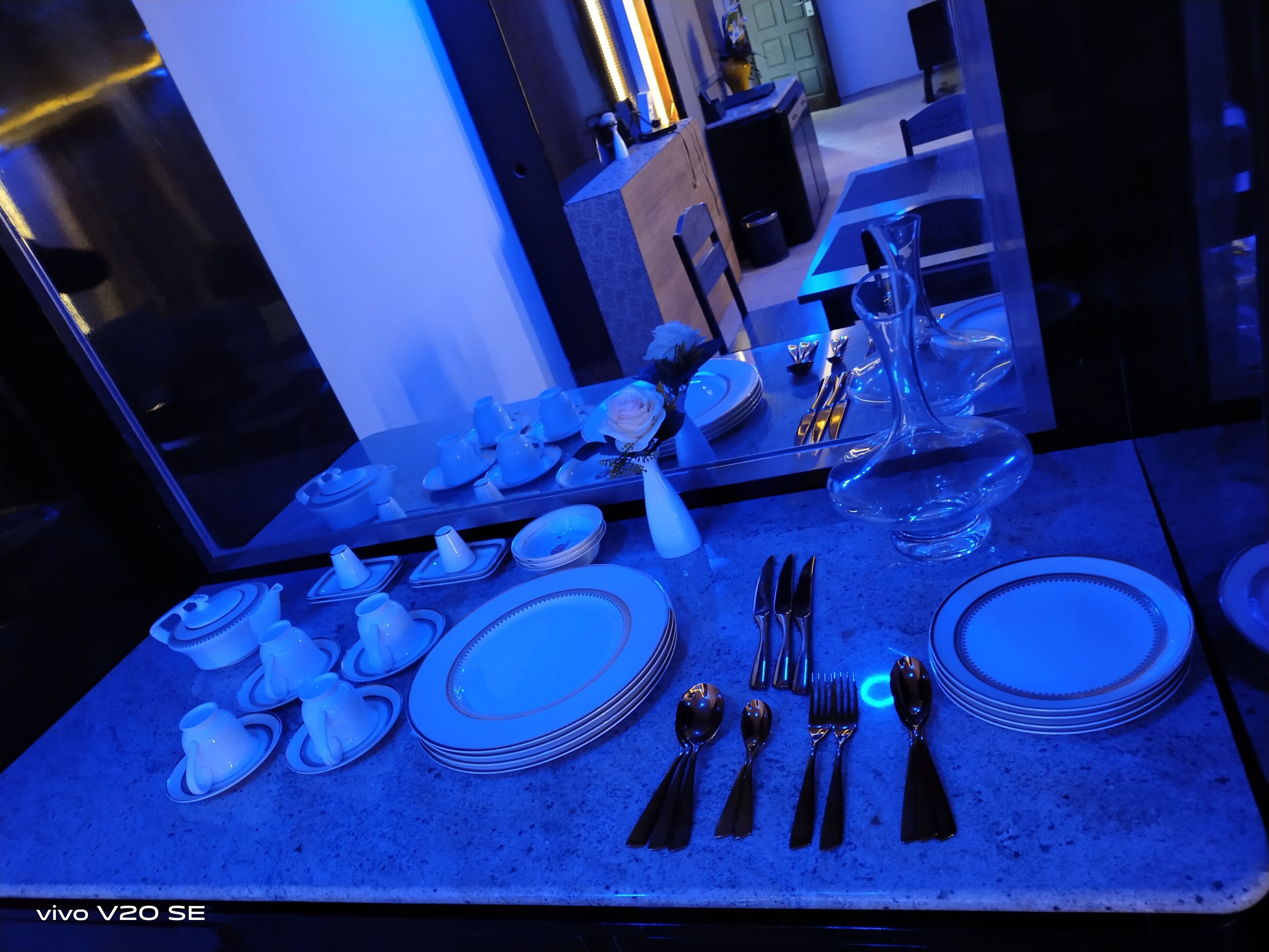 Mesa de jantar posta com pratos, tigelas, xícaras e talheres, refletindo uma luz azul.