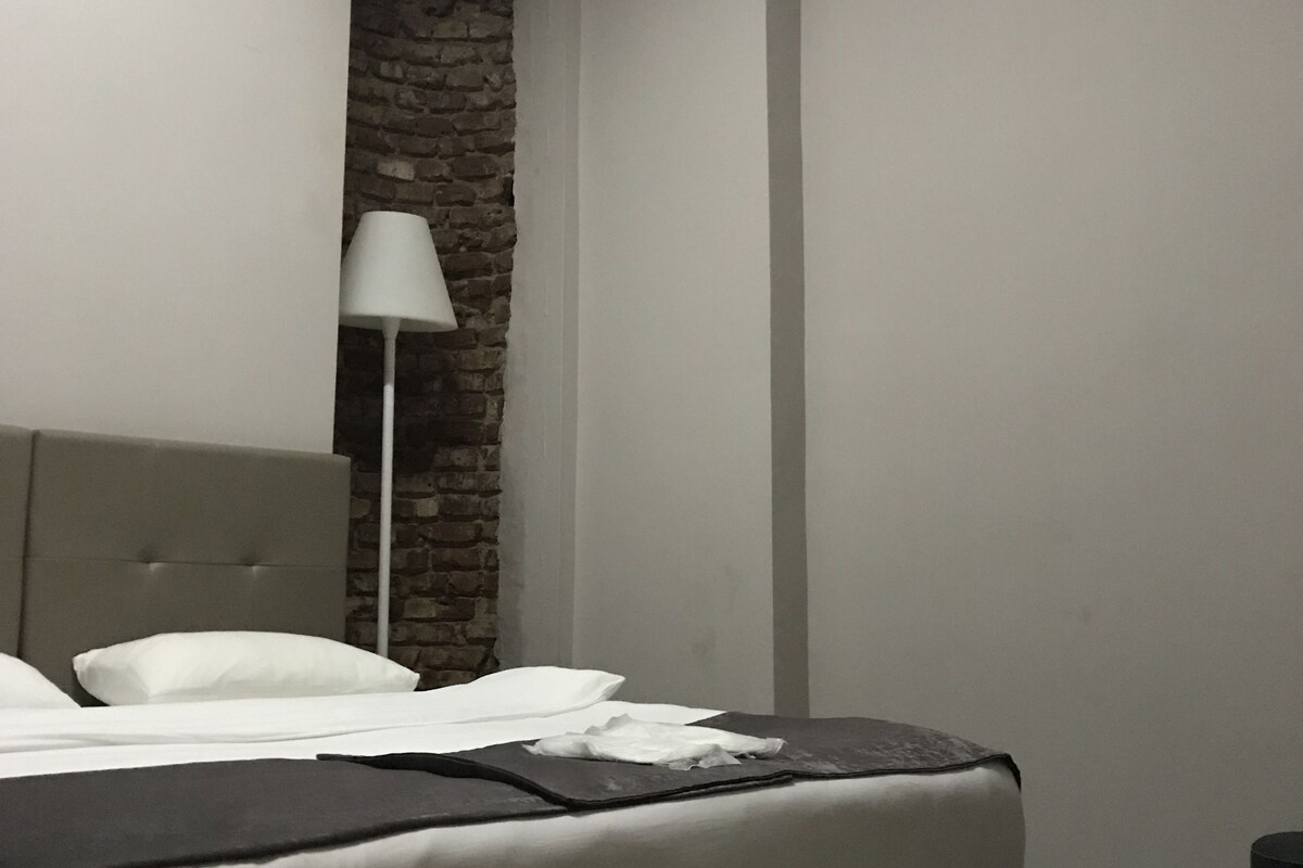 Cama bem arrumada com lençóis brancos e cabeceira cinza, abajur branco e parede de pedra decorativa.