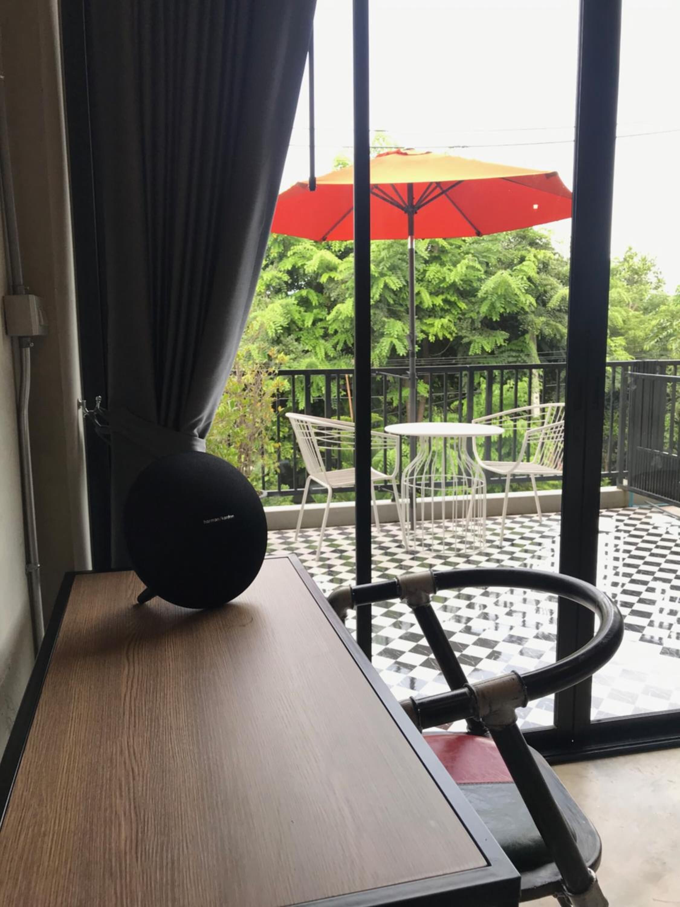 Um quarto com chão xadrez, uma mesa com um objeto preto, uma cadeira e vista para área externa com um guarda-sol vermelho e vegetação.
