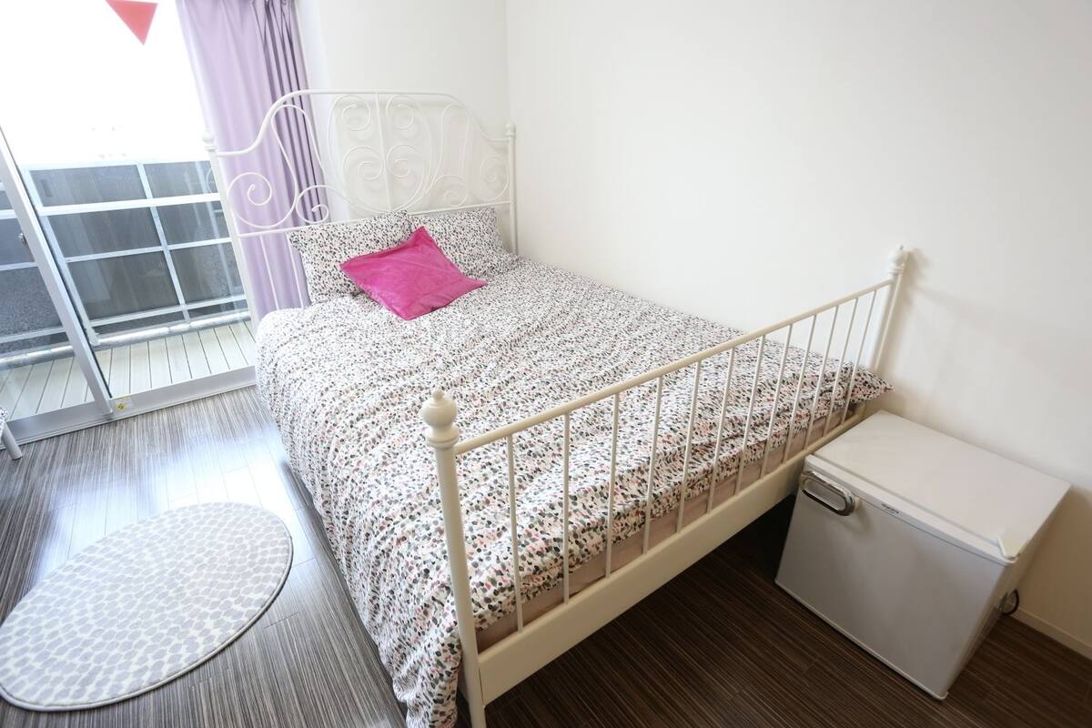 Quarto individual com uma cama de estrutura metálica, uma mesinha de cabeceira, uma cadeira e uma janela com cortinas.