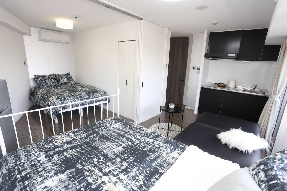 Quarto compacto com cama, mesa pequena, guarda-roupa e área de cozinha compacta.