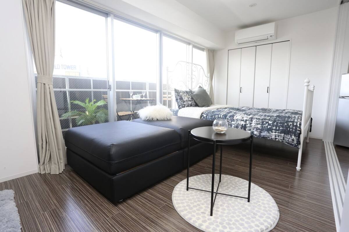 Quarto de hotel moderno com uma janela grande, uma cama com roupa de cama estampada, um banquinho preto, uma mesinha redonda pequena e um radiador branco.