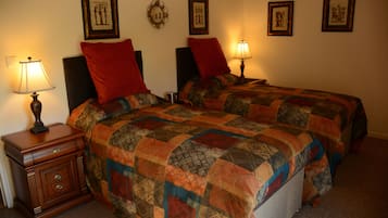 Quarto com duas camas, cada uma com travesseiro vermelho e colcha estampada. Há duas mesas de cabeceira com abajures, e as paredes são decoradas com quadros emoldurados.