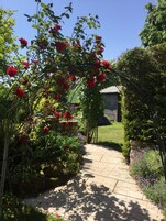 Um caminho no jardim com um arco de rosas, muros de pedra e diversas plantas.