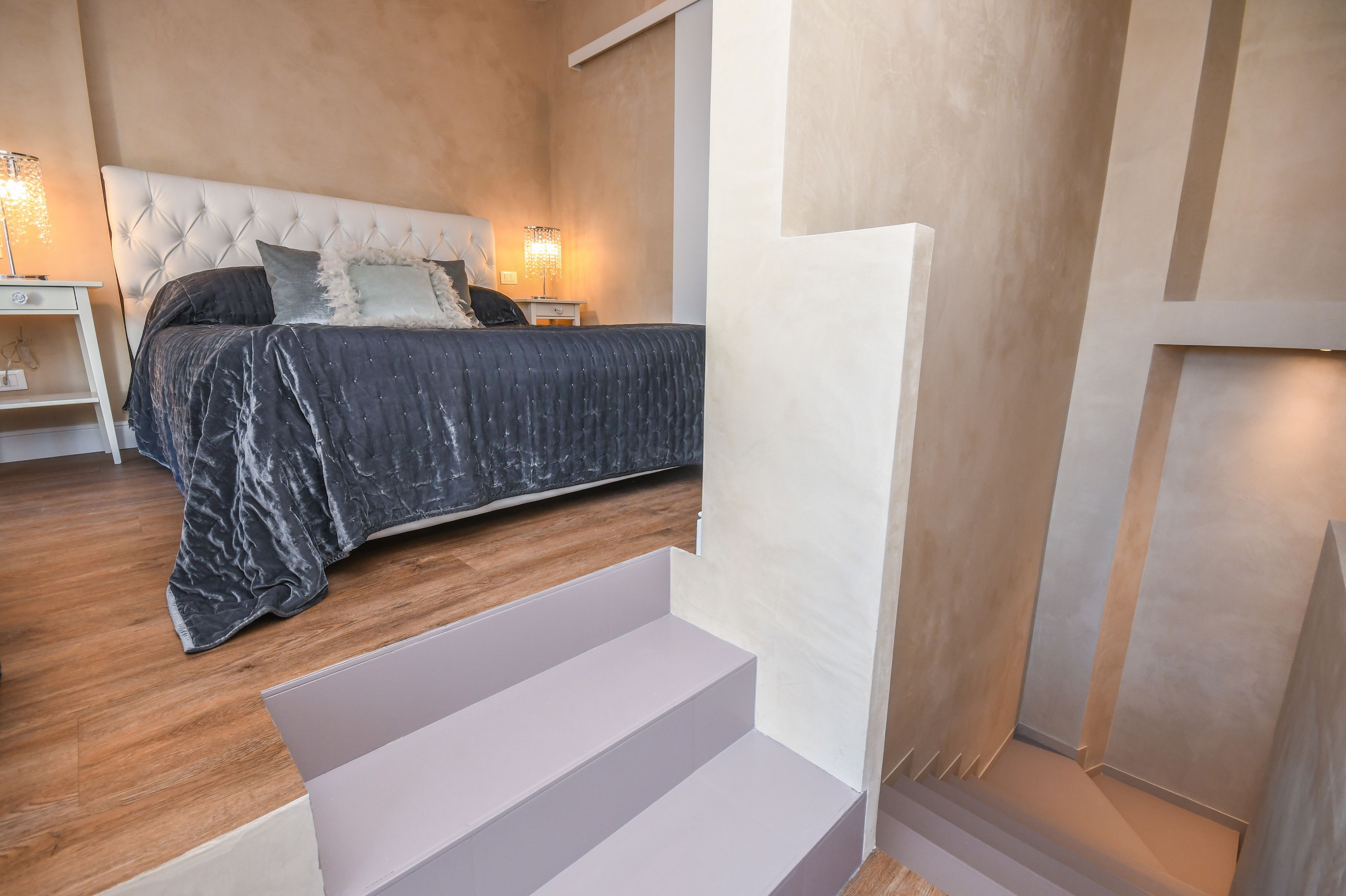 Quarto com cabeceira estofada, cama com colcha estampada escura, mesa de cabeceira com luminária e piso de madeira.