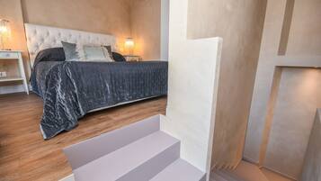 Quarto com cabeceira estofada, cama com colcha estampada escura, mesa de cabeceira com luminária e piso de madeira.