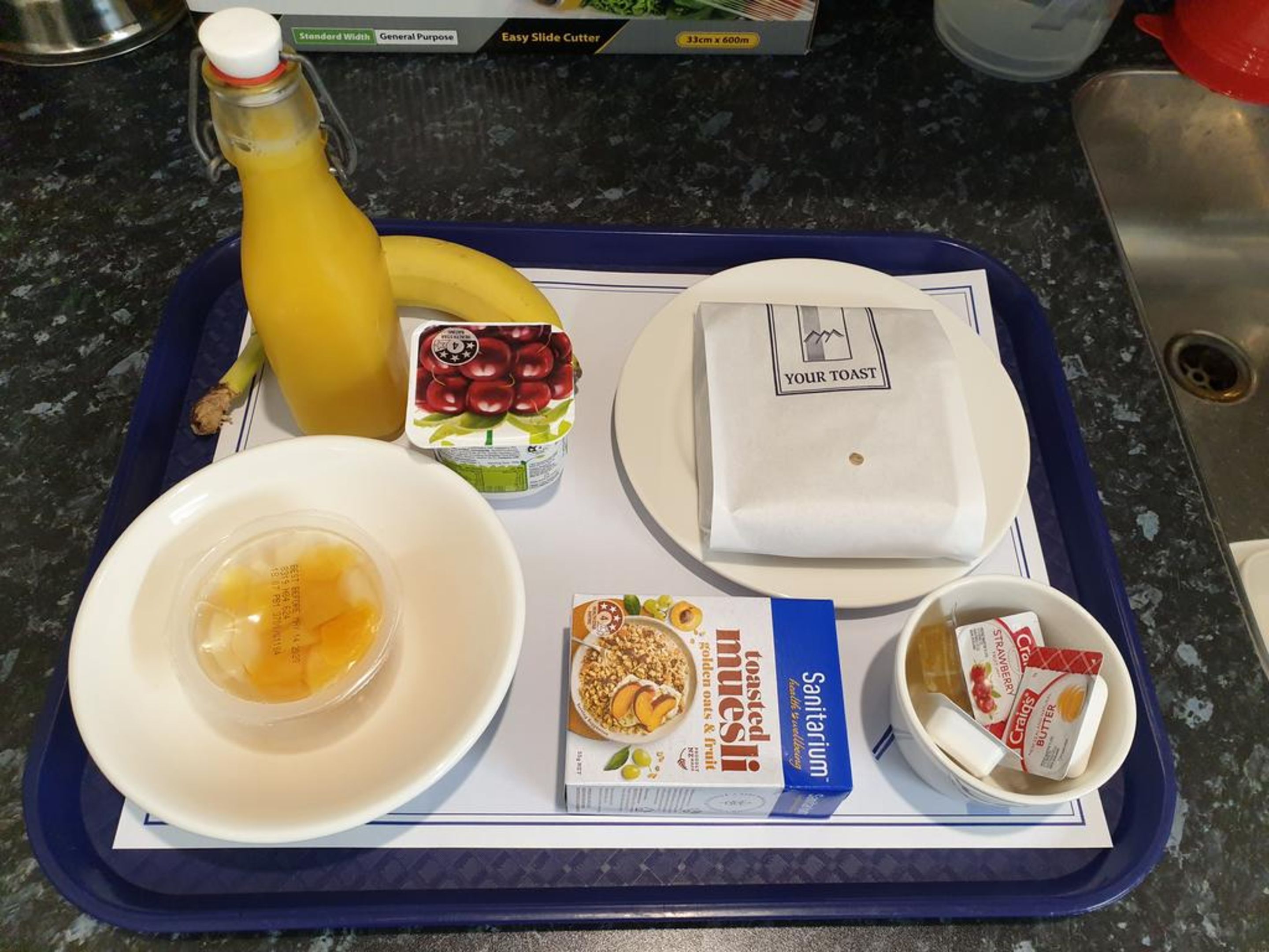 Uma bandeja com uma banana, um recipiente com cerejas, uma tigela de cereal, uma embalagem de torradas e uma pequena xícara de manteiga.