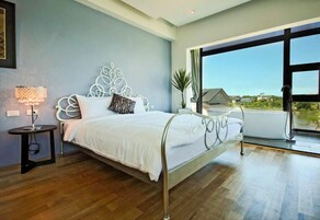 Quarto com estrutura de cama de metal, mesa de cabeceira com abajur e uma grande janela com vista para o exterior.