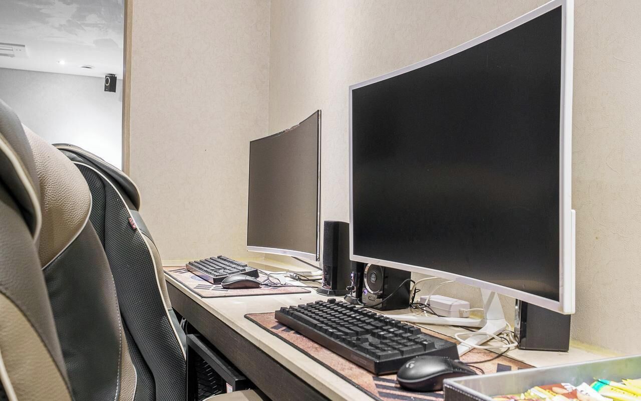 Quarto de hotel com escrivaninha, dois monitores, teclado, rato e cadeira.