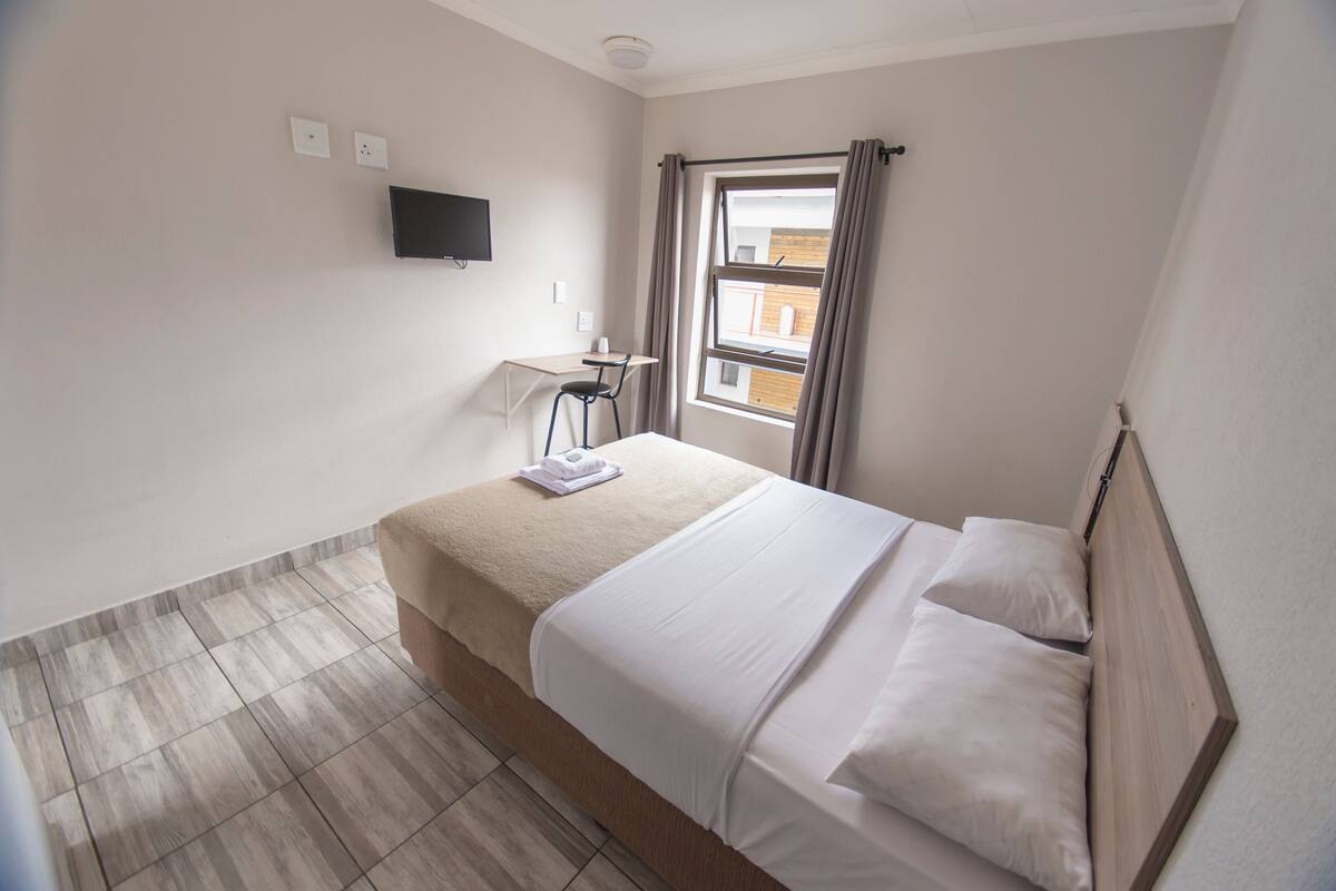 Quarto de hotel com cama, escrivaninha, cadeira, televisão fixada na parede e janela com cortinas.