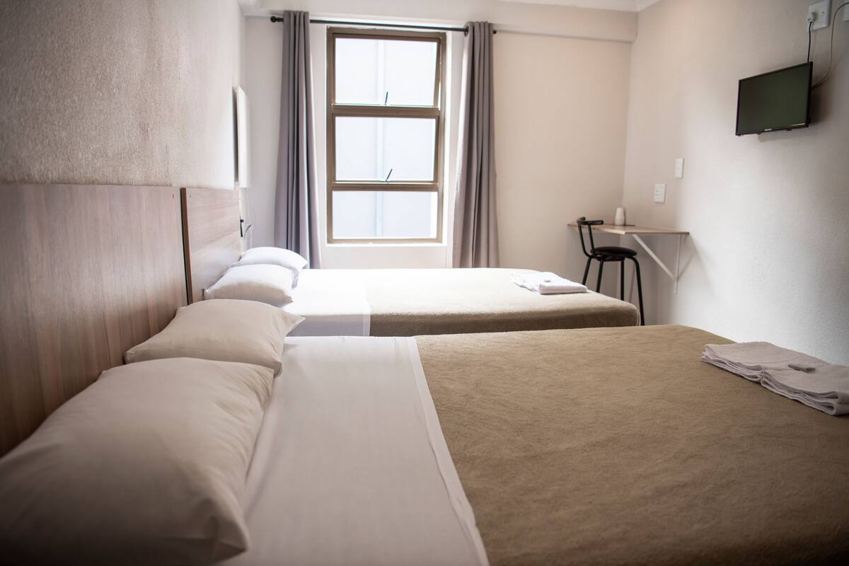 Quarto de hotel com duas camas, uma escrivaninha, uma cadeira, uma televisão e uma janela com cortinas.