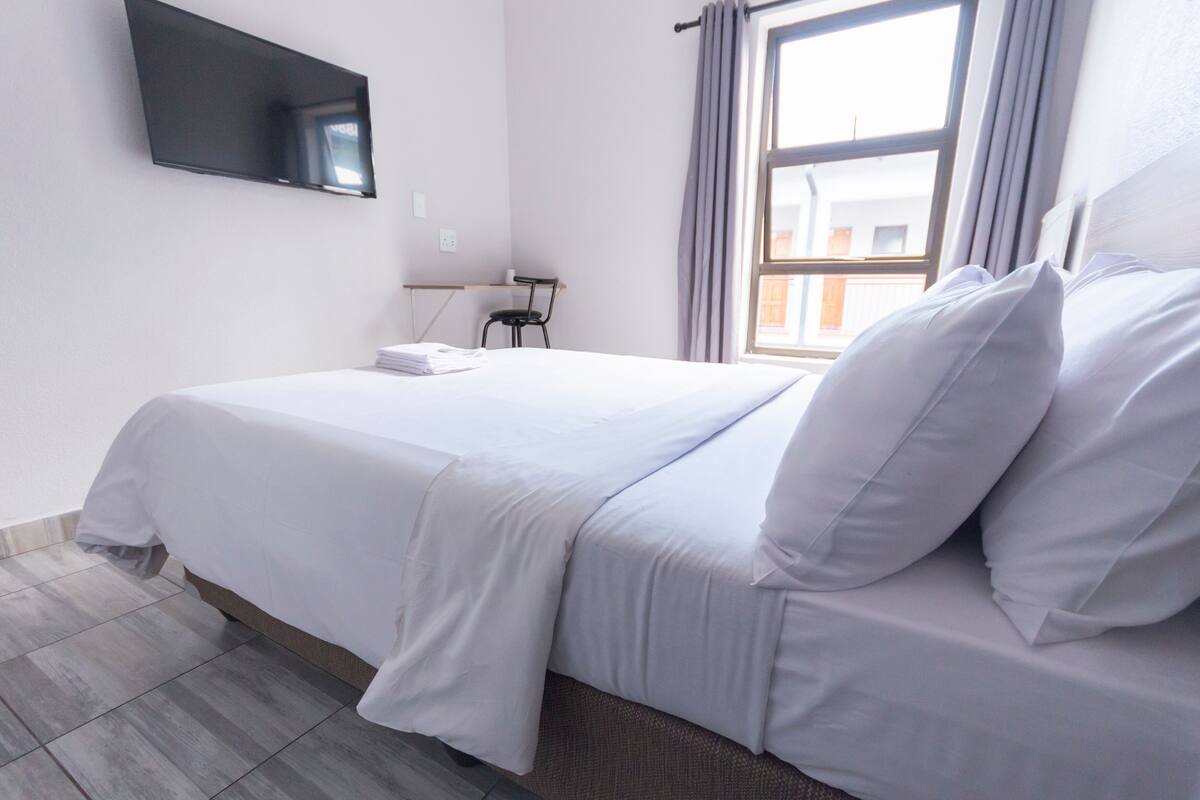 Quarto de hotel com cama, televisão de tela plana na parede, escrivaninha com cadeira e janela com cortinas.