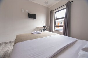 Quarto de hotel com cama, escrivaninha, cadeira, televisão na parede e janela com cortinas.
