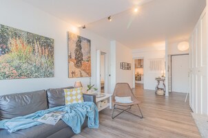 Uma sala de estar moderna com um sofá de couro, uma mesa de centro e uma poltrona. Há duas pinturas na parede e uma porta que dá para outro cômodo.
