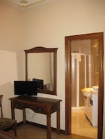 Quarto com escrivaninha de madeira, televisão, cadeira, espelho e banheiro visível por uma porta aberta.