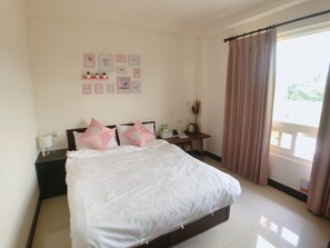 Uma cama bem arrumada com edredom branco e almofadas decorativas cor-de-rosa, uma mesinha de cabeceira com abajur e uma pequena planta, e um painel decorado com quadros.