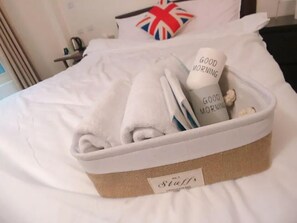 Cama bem arrumada com manta branca, toalha dobrada e um recipiente com a etiqueta "Bom Dia" contendo produtos de higiene pessoal.