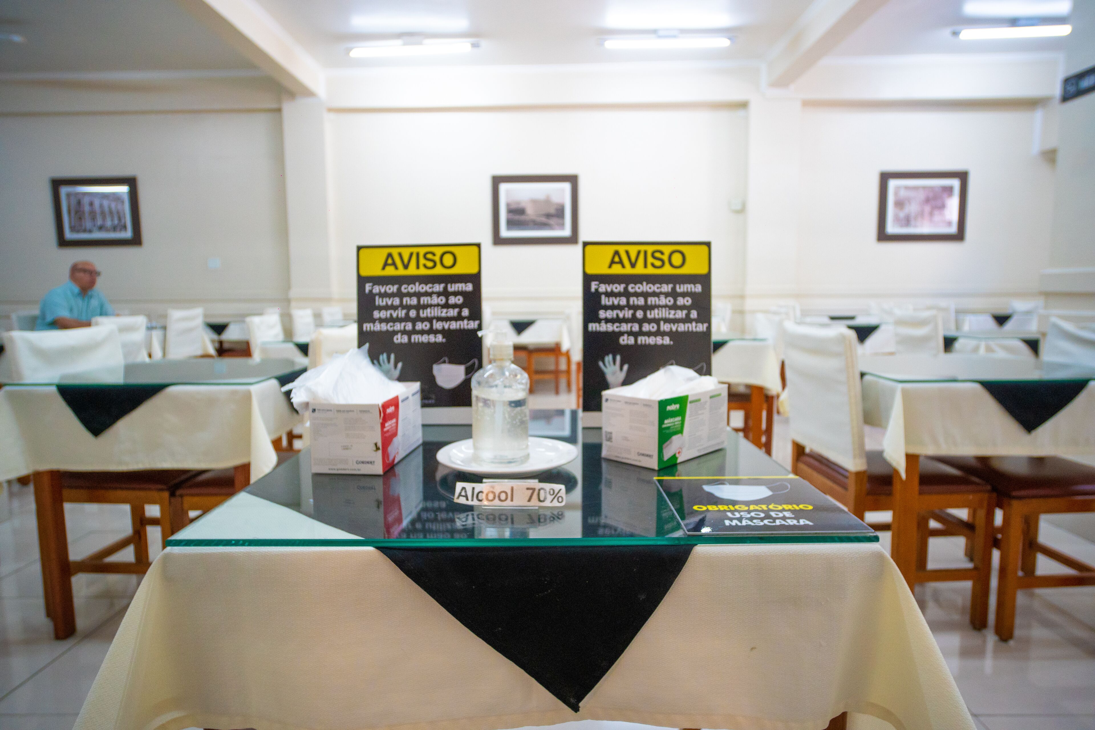 Sala de conferência com mesas organizadas para uma reunião. Há placas nas mesas instruindo os participantes a usarem máscaras. Uma garrafa de álcool e uma caixa de máscaras estão na mesa.