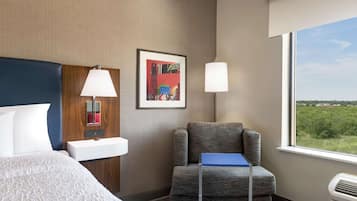 Quarto de hotel com uma cama, uma escrivaninha com luminária, uma cadeira e uma janela com vista para o verde.